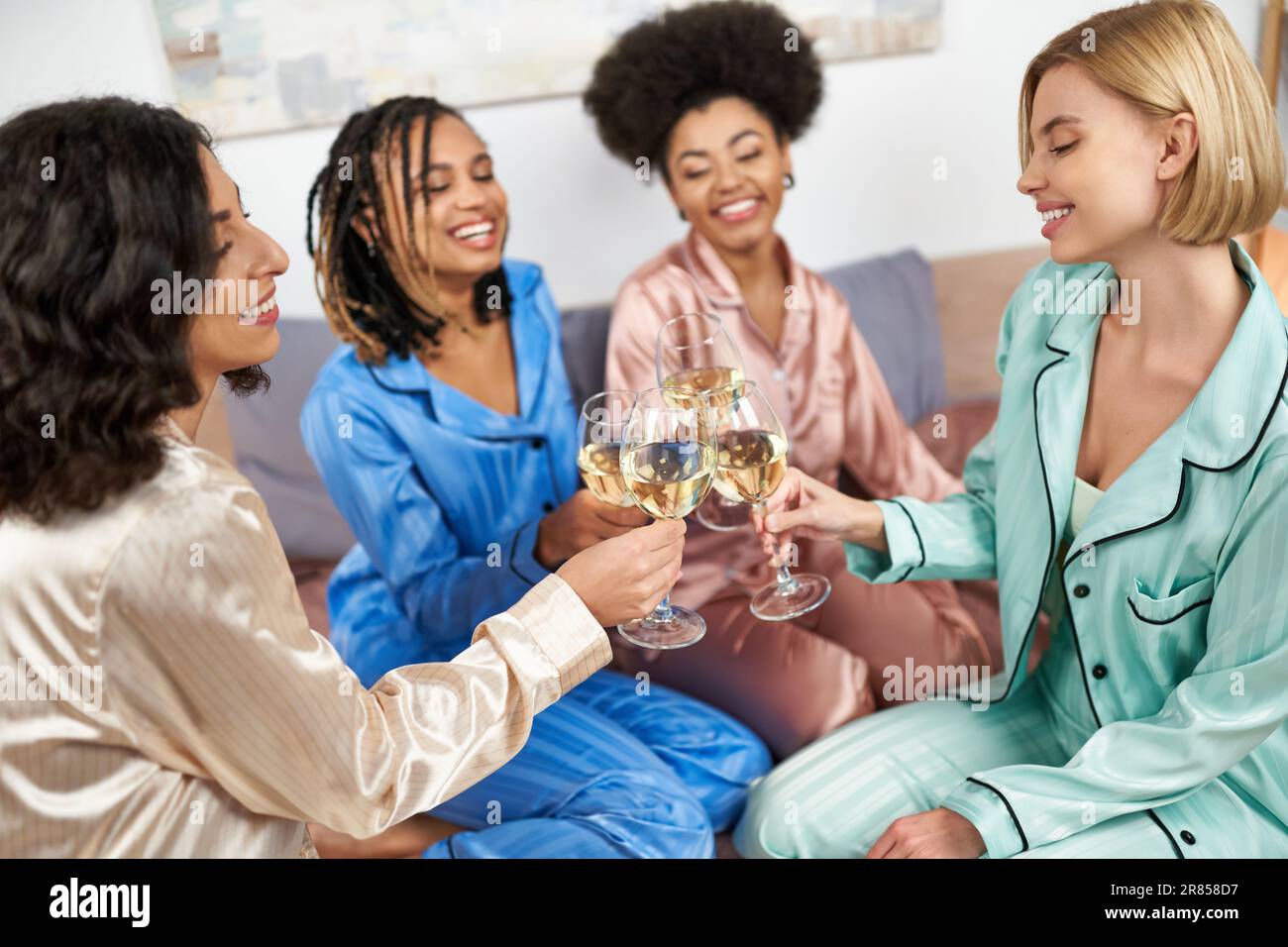 Smiling multiethnic women in colorful pajama toasting with glasses of ...
