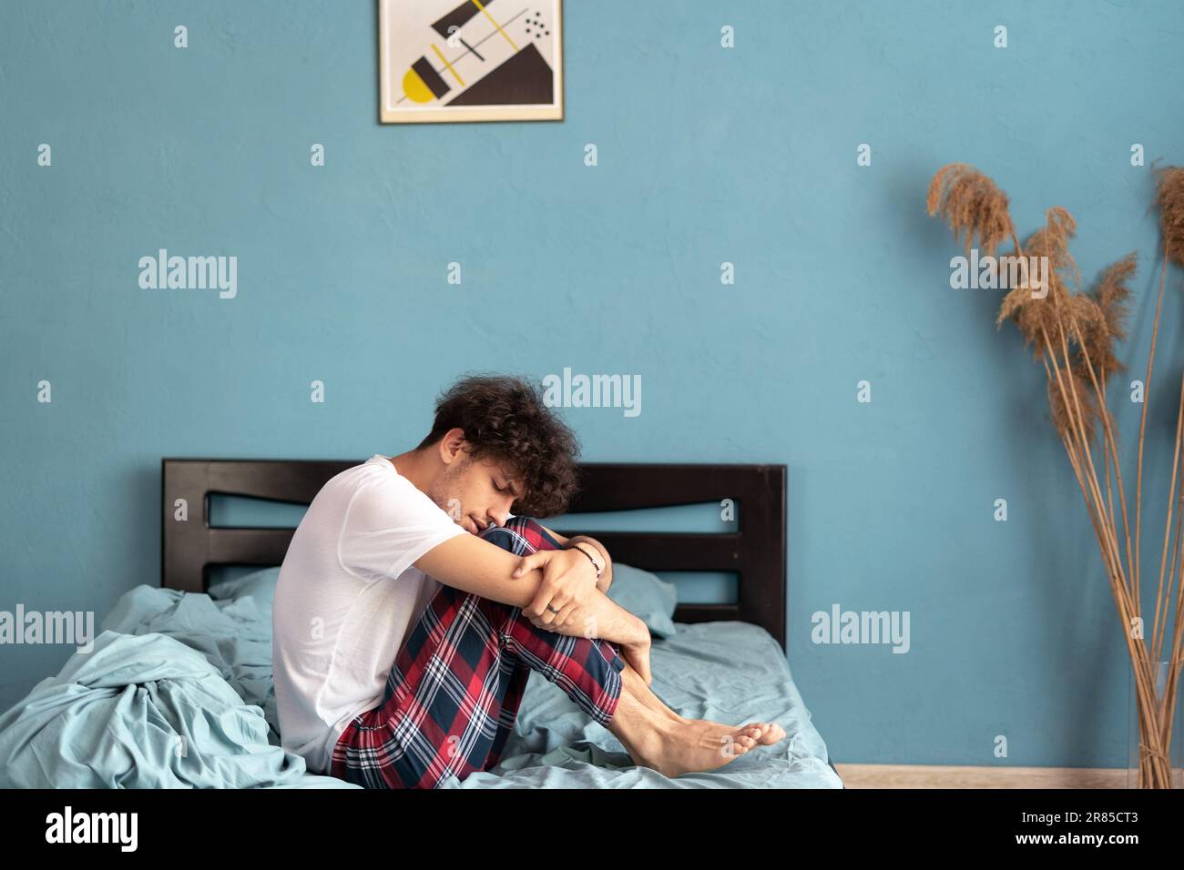 Young man suffering from depression sits on the bed hugging his legs ...