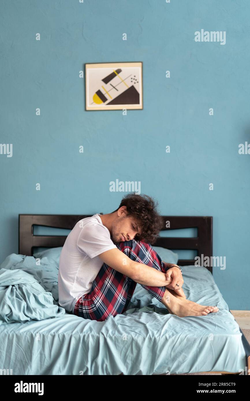 Sad young man suffering from depression sitting on bed in pajamas ...