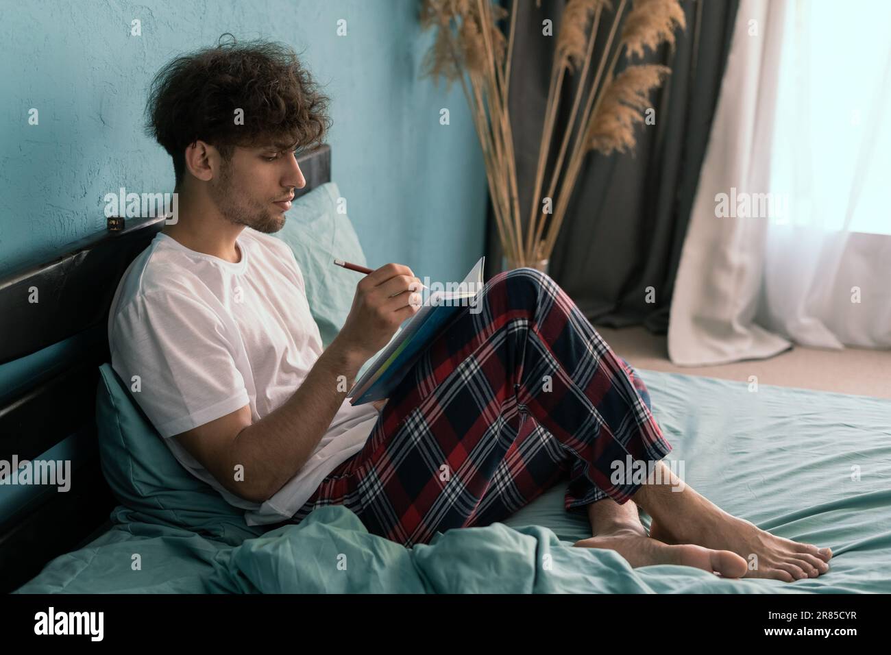 Handsome young man taking notes in notebook while lying on bed writing ...