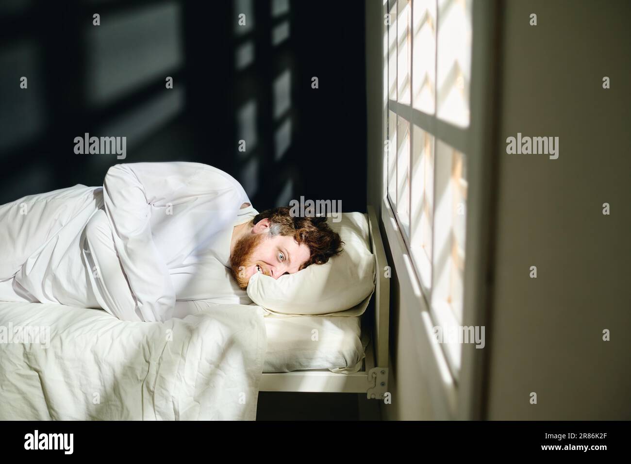 Young deranged man in straitjacket biting pillow while lying on bed by ...