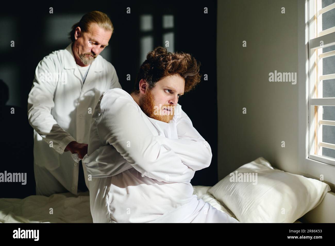 Mature psychiatrist standing by bed of insane male patient suffering ...