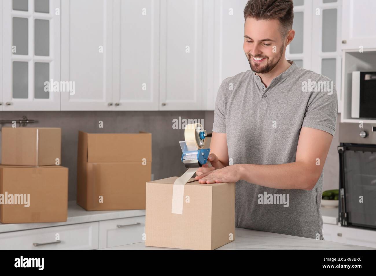 Smiling man taping box with adhesive tape dispenser in kitchen, space ...