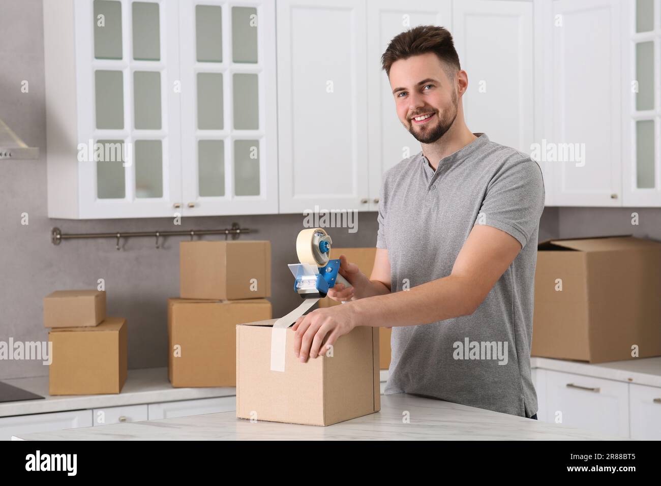 Smiling man taping box with adhesive tape dispenser in kitchen, space ...