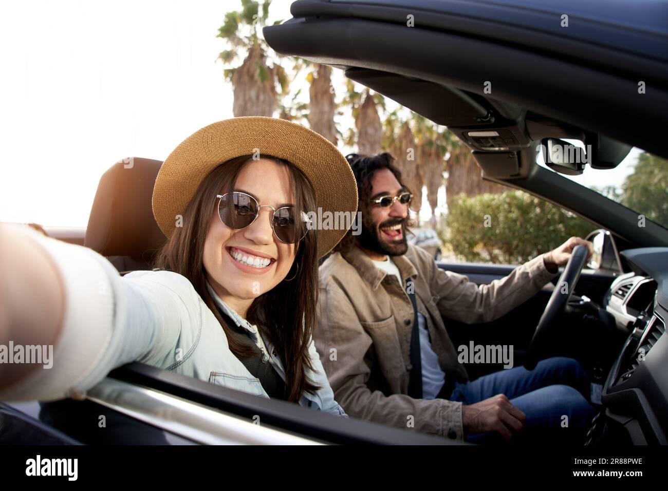 Caucasian young couple driving a cabrio car. Woman takes selfie with ...