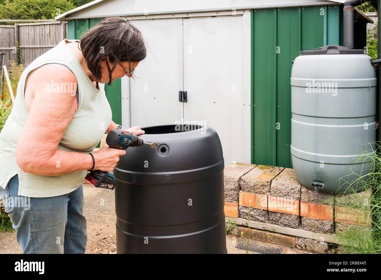 Woman fitting a second rainwater butt in her garden to take overflow ...