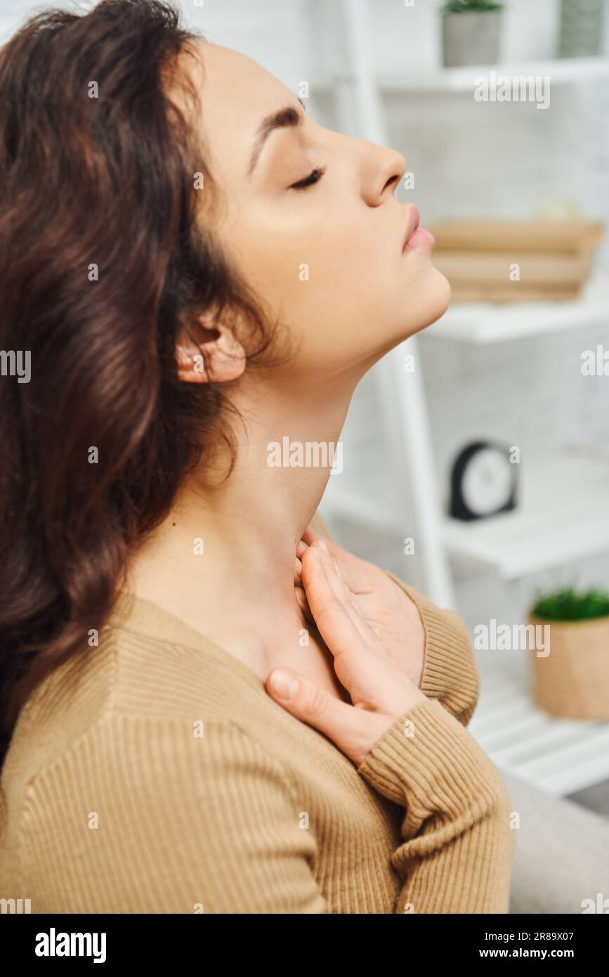 Side view of young brunette woman in casual jumper touching neck during ...