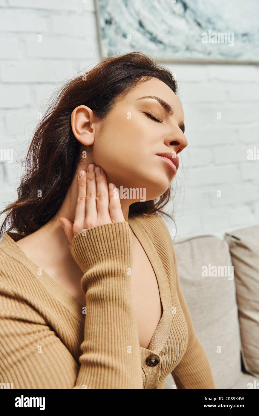 Portrait of young brunette woman in casual jumper massaging neck and ...