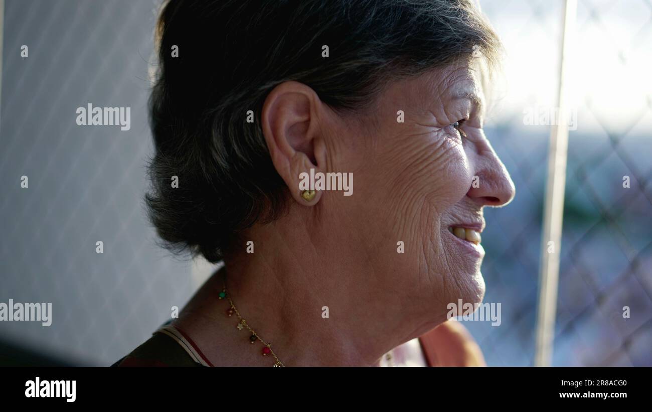 Contemplative happy older woman standing by window looking outside ...