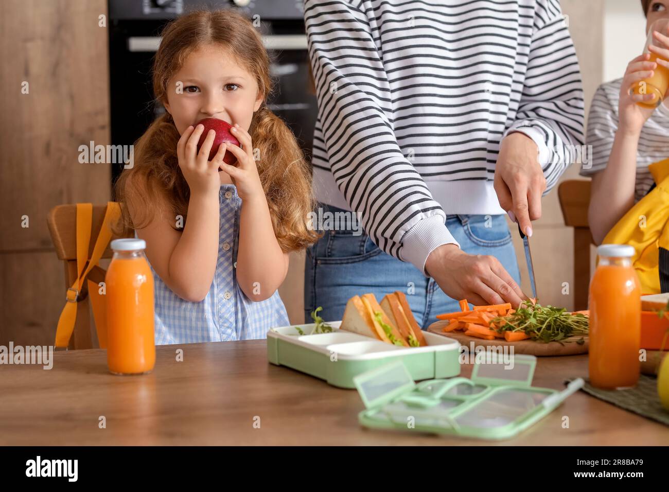 Little girl eating apple while her mother preparing school lunch in ...