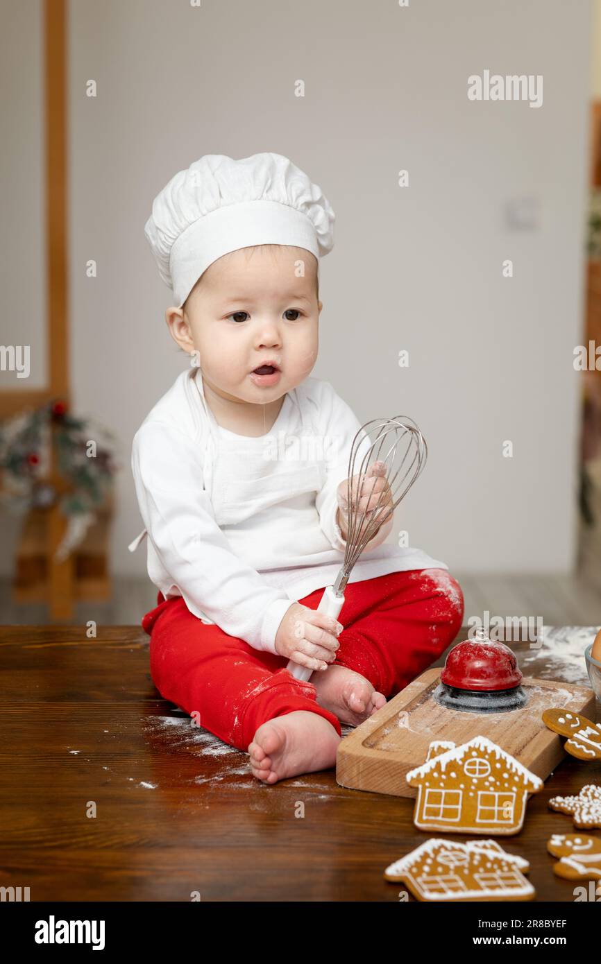 An Asian baby in a chef's cap and apron sitting on the kitchen table ...