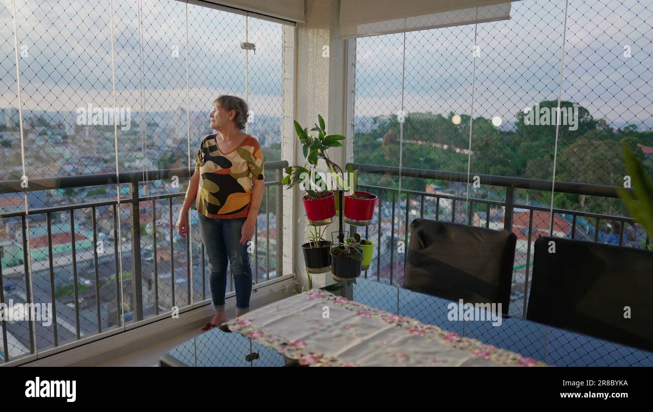 Contemplative senior woman standing at apartment balcony looking at ...