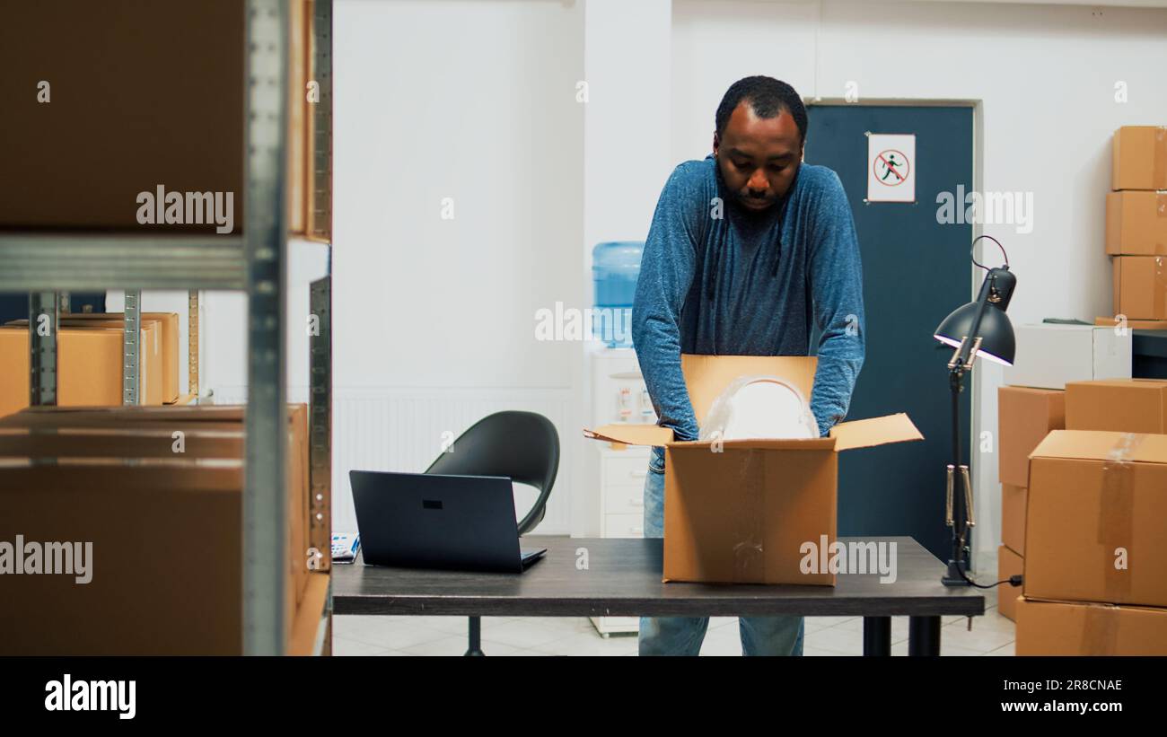 Male warehouse worker arranging supplies in packages, using laptop to ...