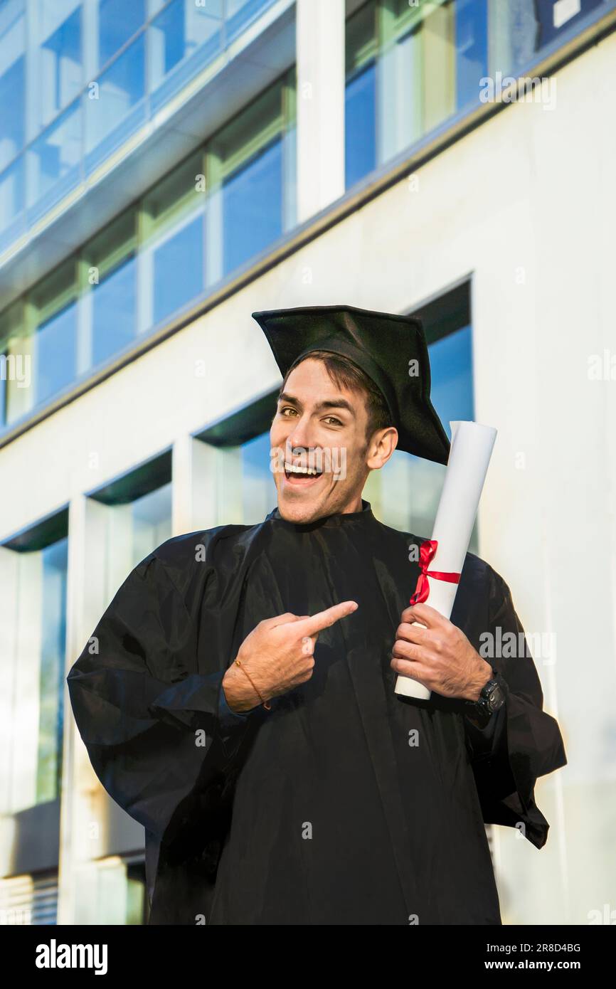 Happy graduated man wearing a bachelor gown and a black mortarboard and pointing at his diploma ...