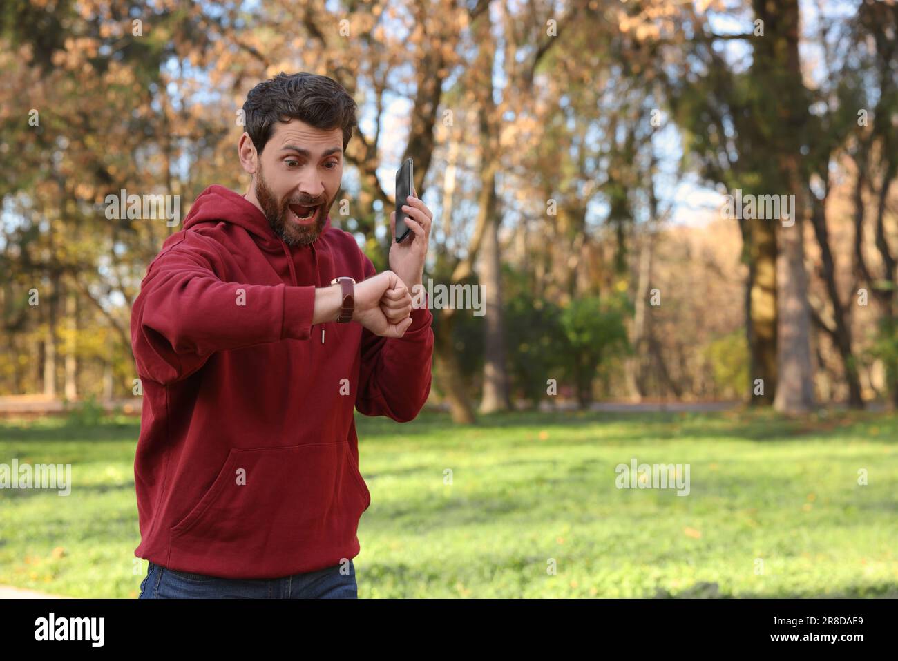Emotional man talking by smartphone and checking time on watch in park ...
