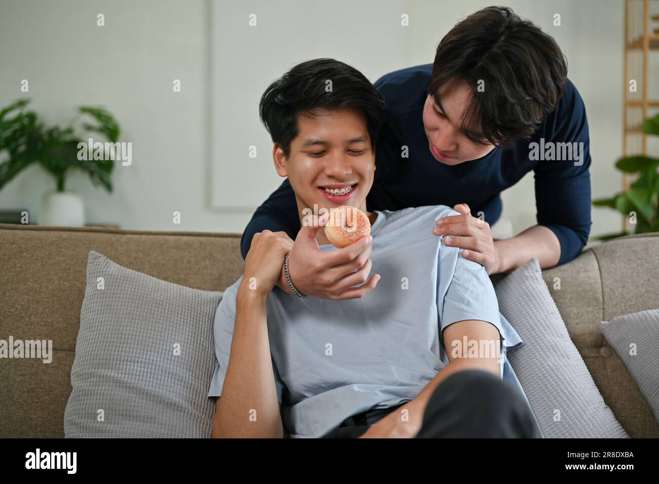 A happy young Asian gay man feeds a doughnut to his boyfriend while ...