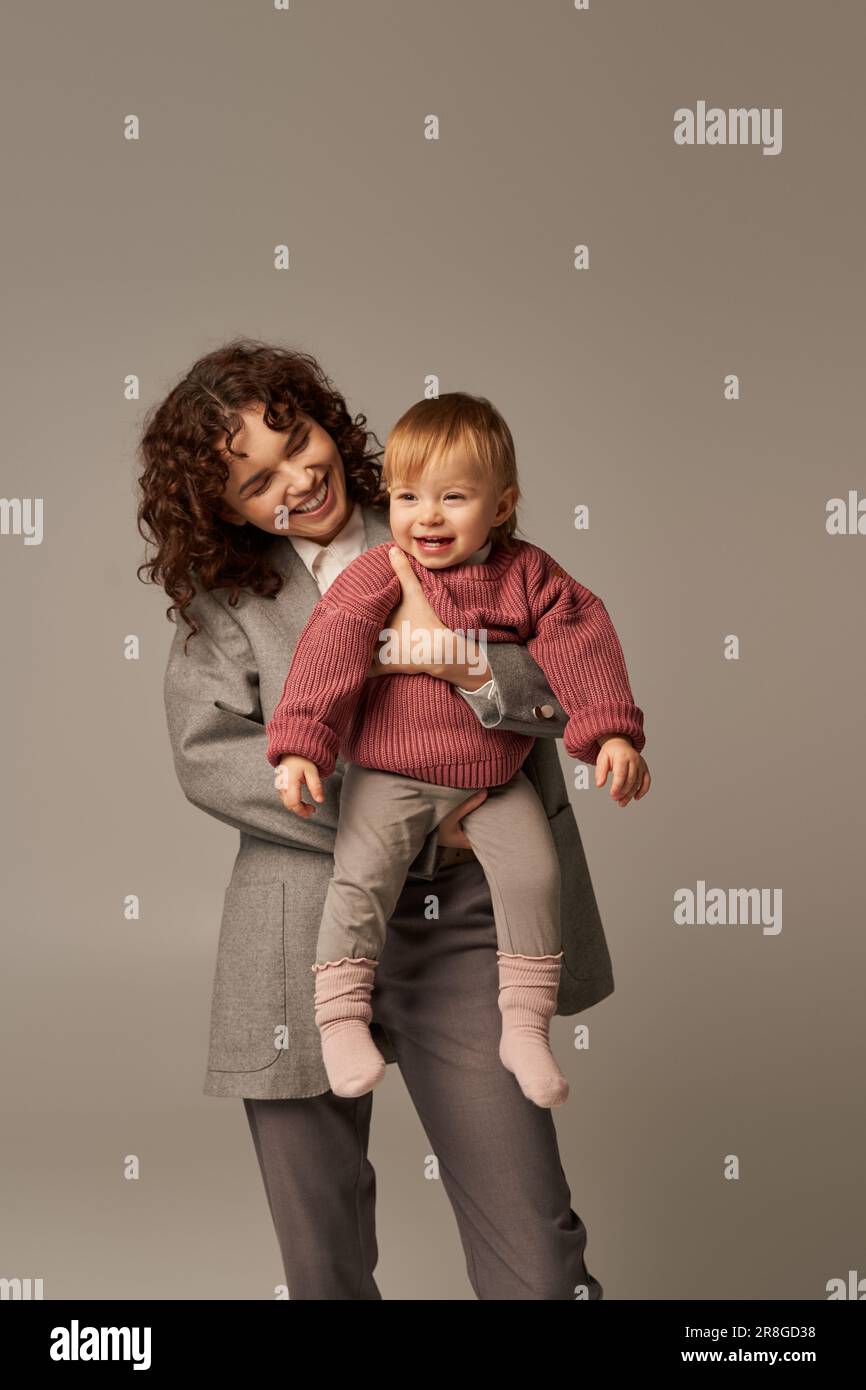 career and family, joyful mother in suit holding smiling child on grey ...