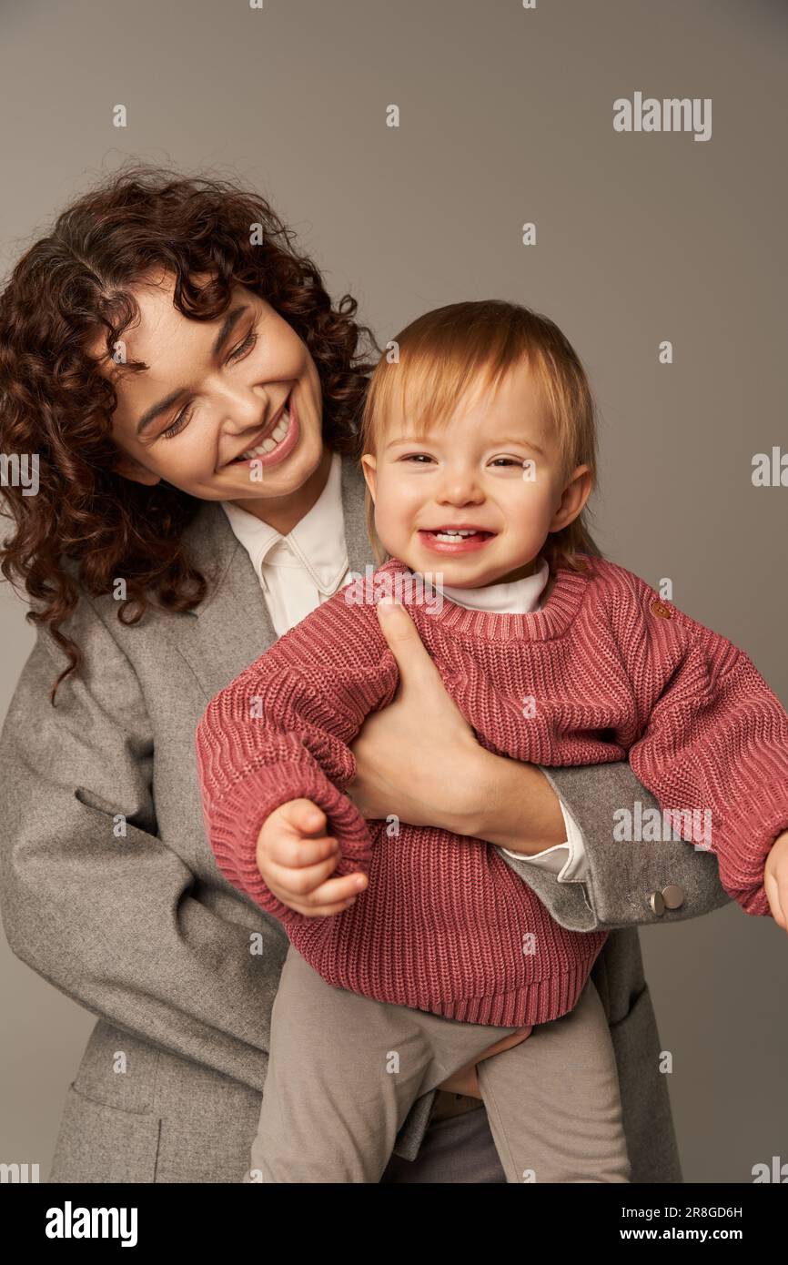 career and family, joyful woman in suit holding happy child on grey ...