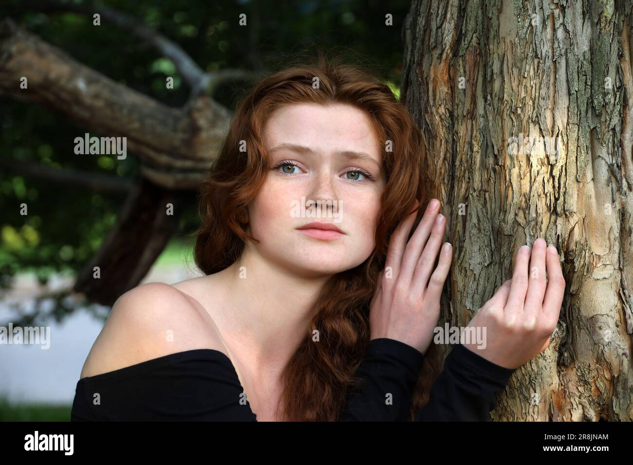 Girl with long red hair and freckles standing near the tree in summer ...