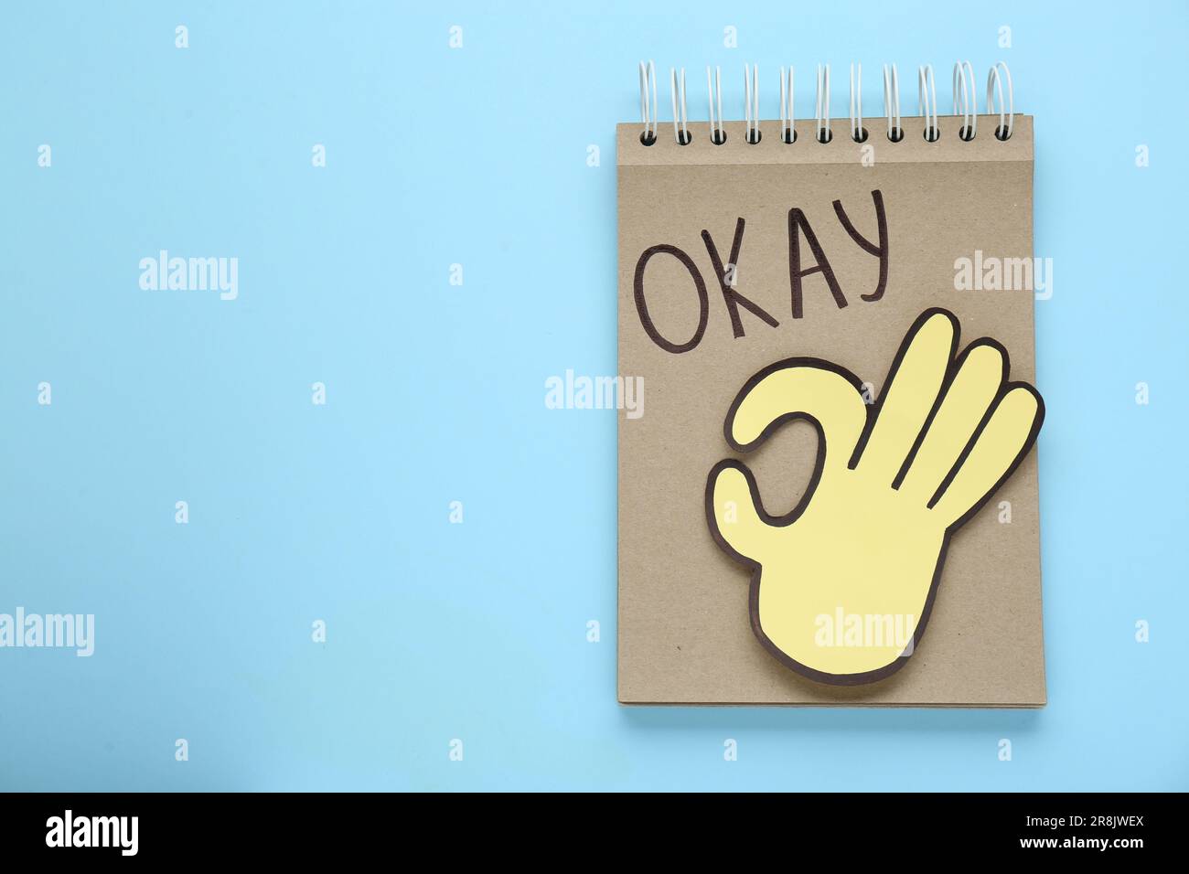 Notepad with word Okay and paper cutout of OK hand gesture on light blue background, top view ...