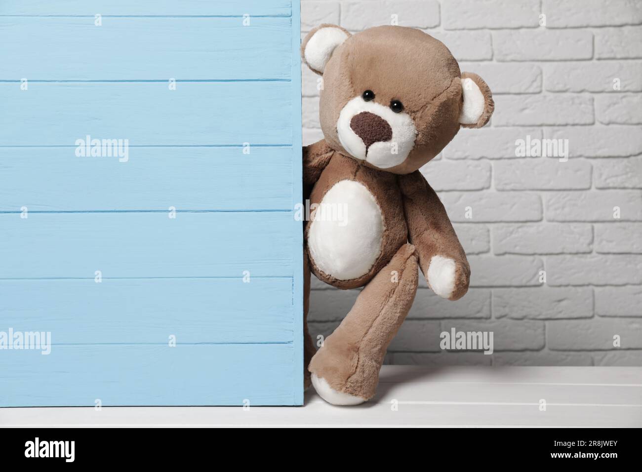 Cute teddy bear peeking out behind light blue wooden wall against brick ...