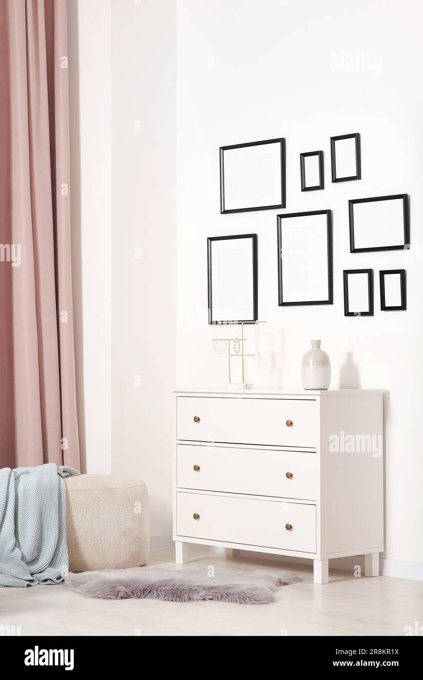 Empty frames hanging on white wall, chest of drawers and ottoman ...