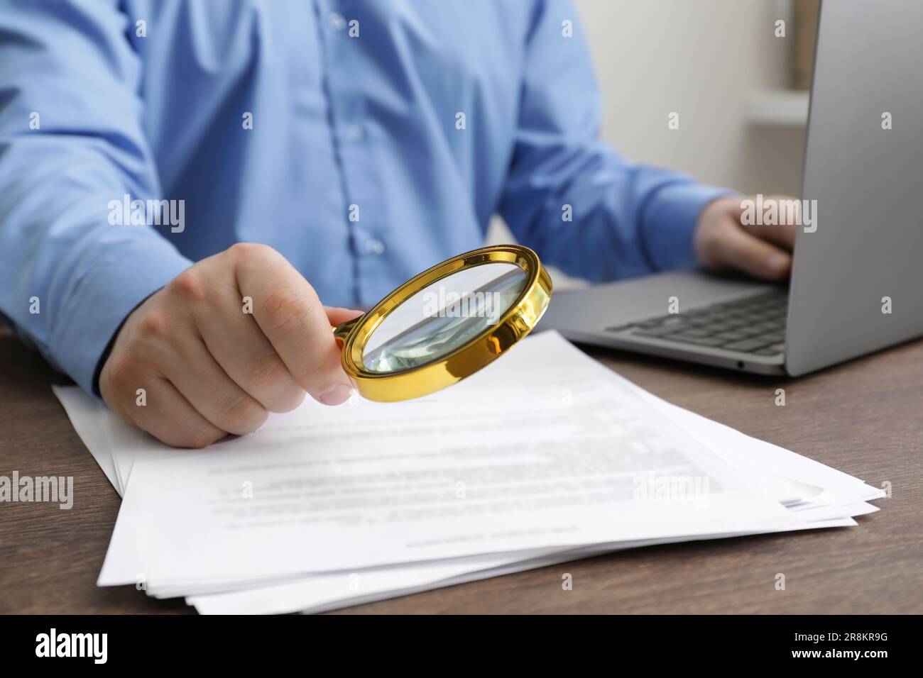 Man holding magnifier near documents while using laptop at wooden table ...