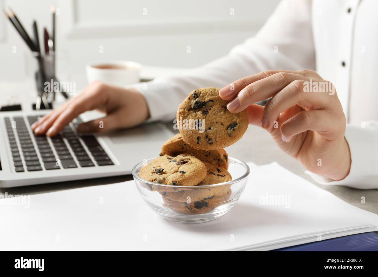 Woman taking chocolate chip cookie from bowl while working with laptop ...