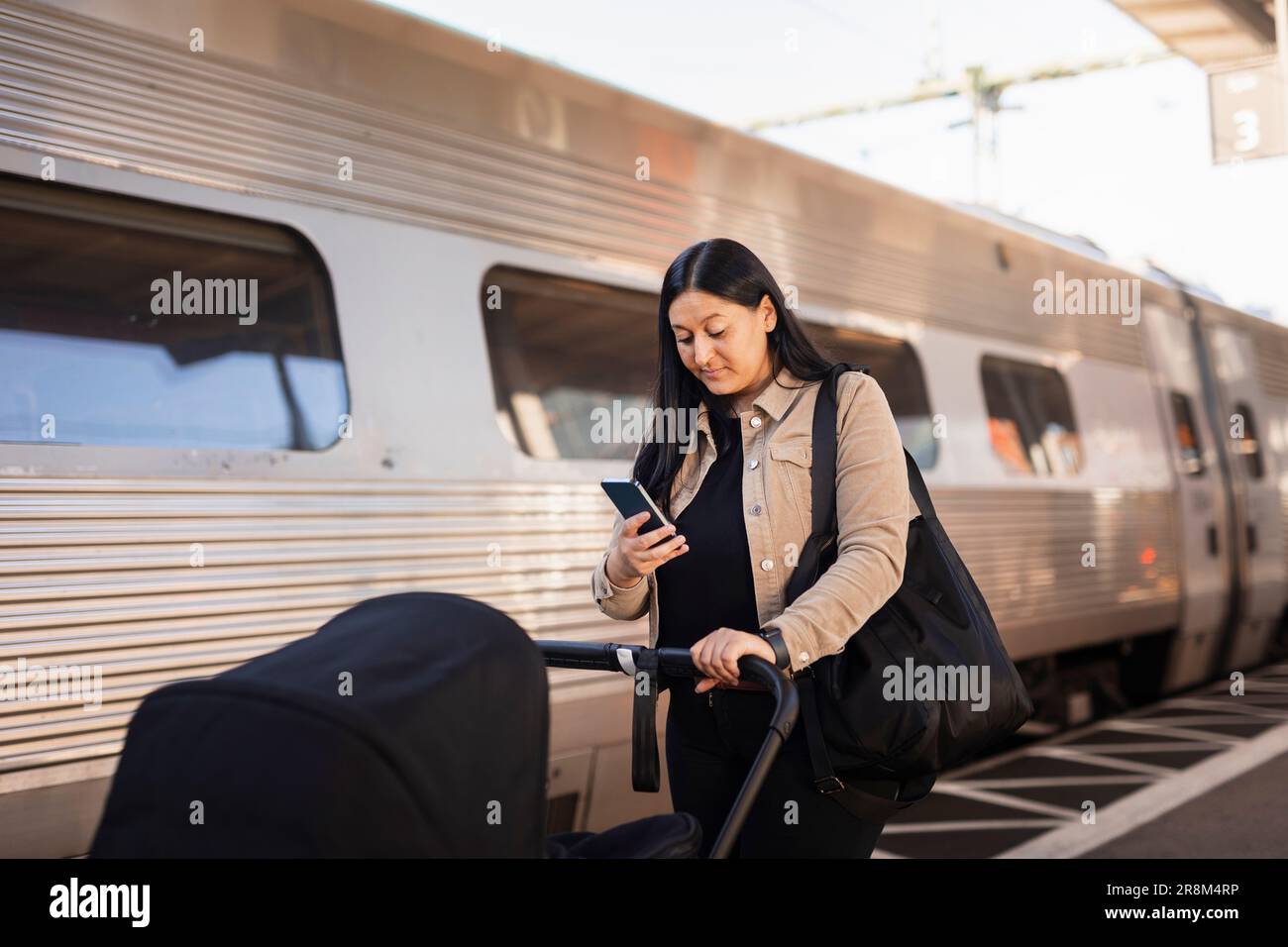 Mid adult woman at train station pushing pram and using cell phone ...