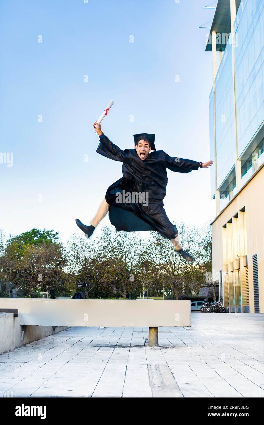 Happy graduated man in a black gown jumping while holding his diploma ...