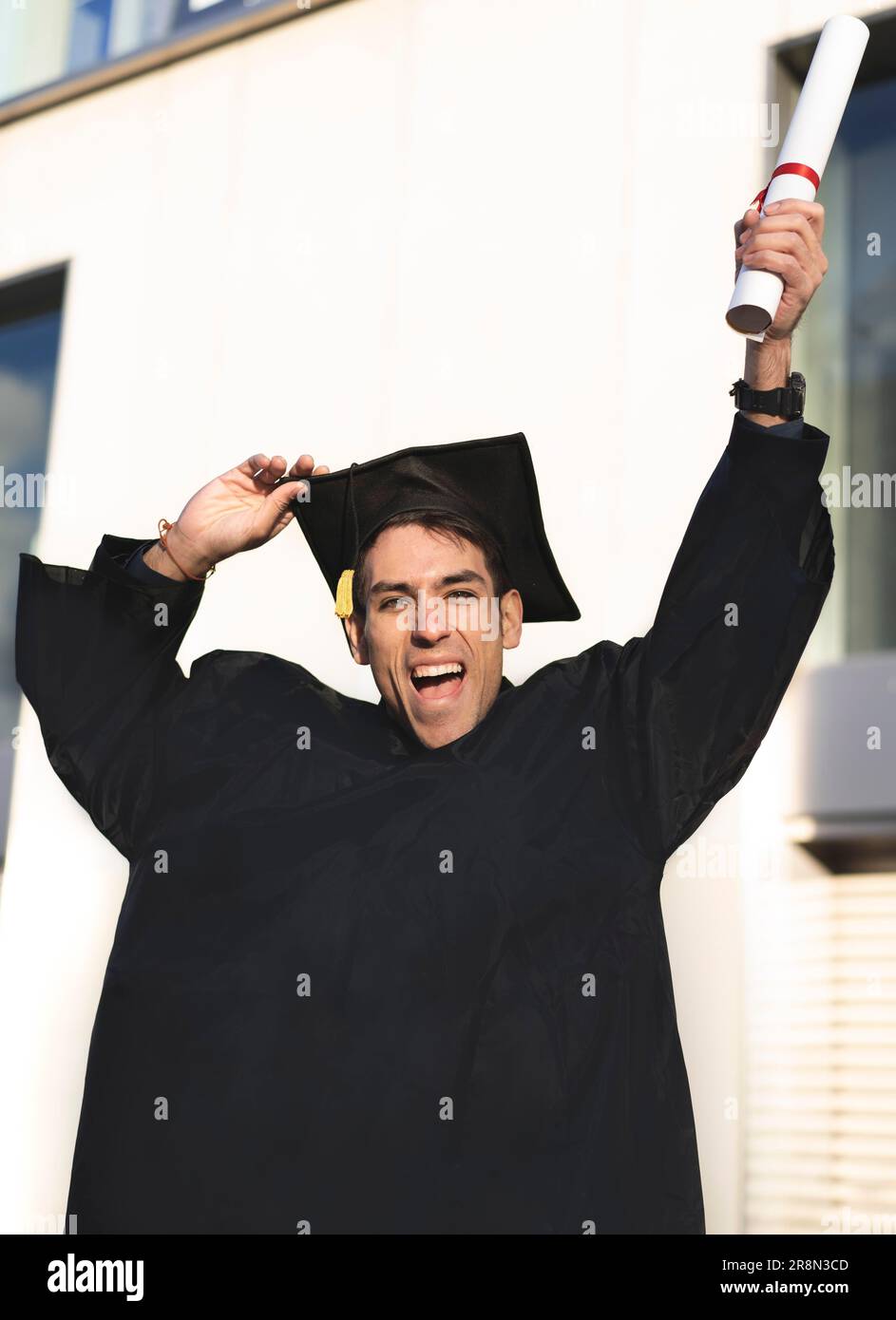 Cheerful smiling graduate man wearing a gown and raising his diploma ...