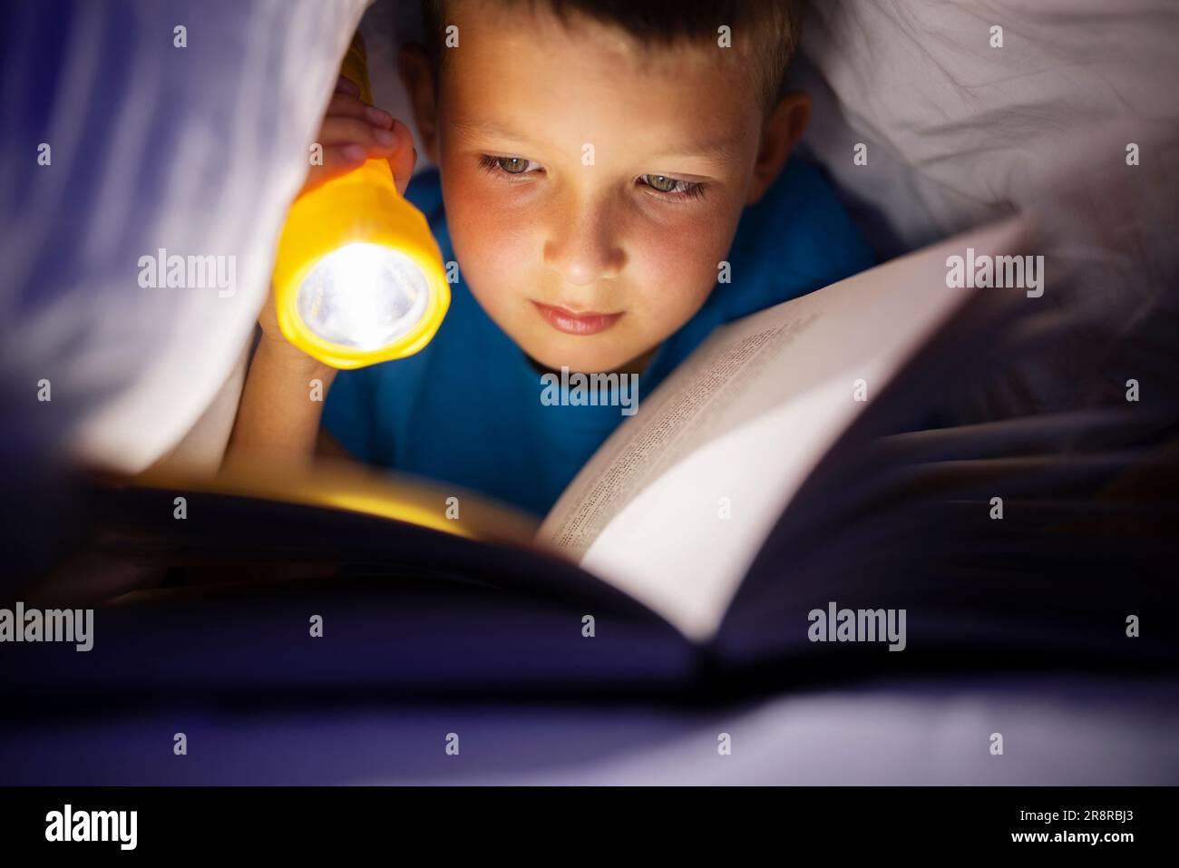 A young boy reading a book under the covers with a flashlight at dark ...