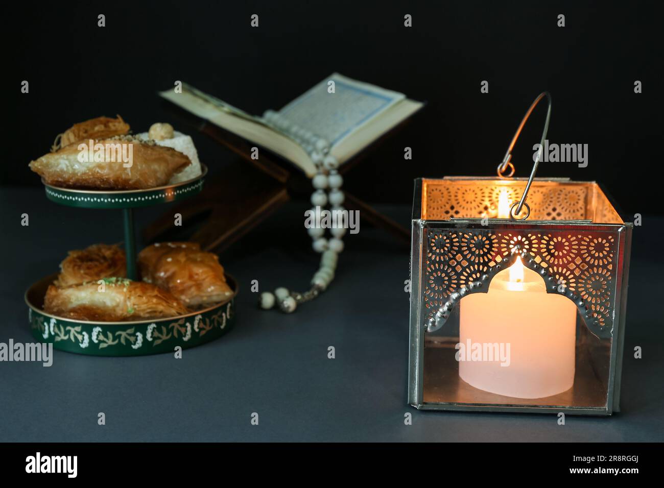 Fanous with burning candle, sweets, Koran and tasbih on dark background ...