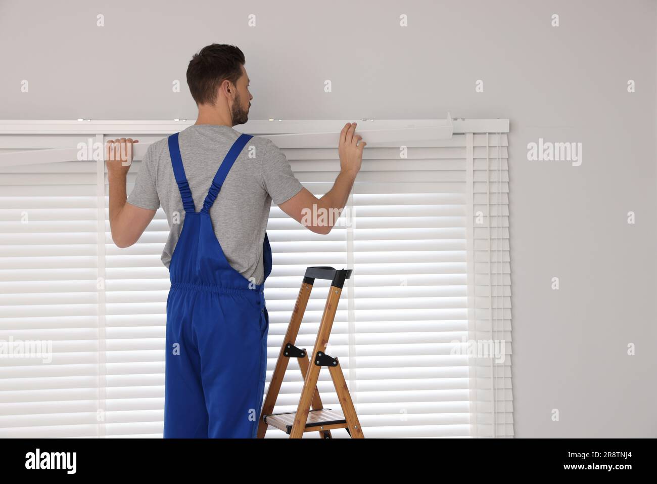 Worker in uniform installing horizontal window blinds on stepladder ...