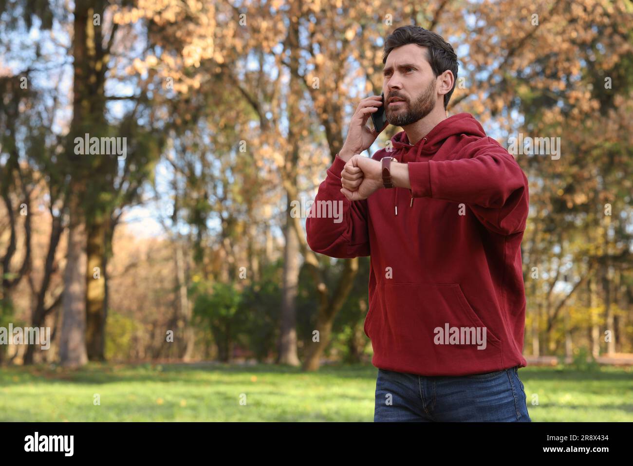 Emotional man talking by smartphone and checking time on watch in park ...