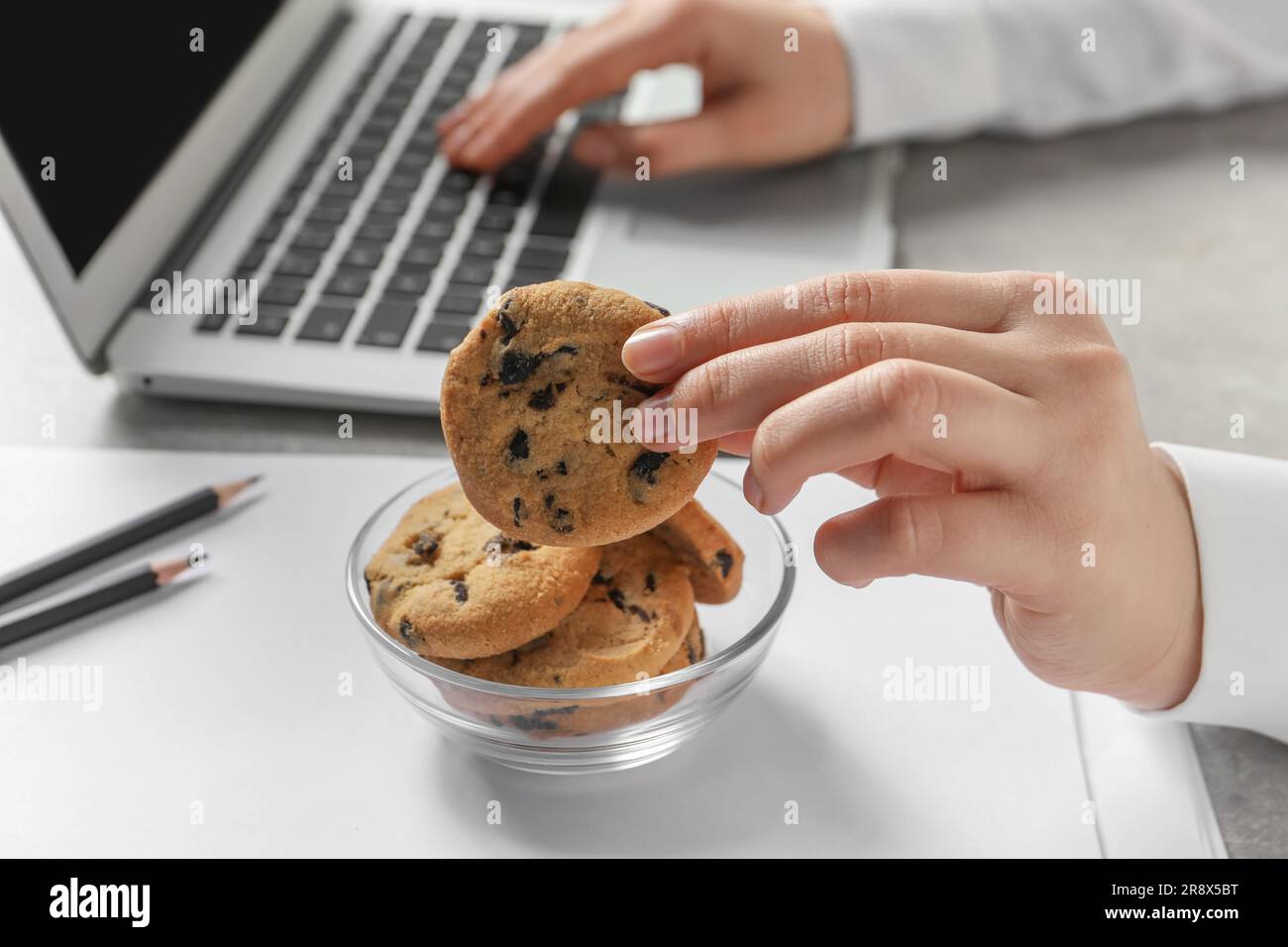 Woman taking chocolate chip cookie from bowl while working with laptop ...