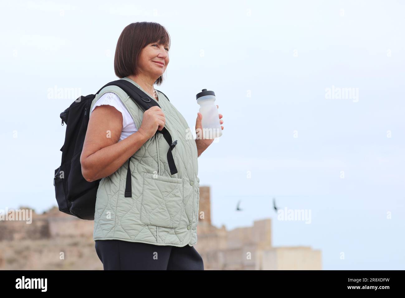 Happy adult woman traveler 50-60 years old wears backpack holds bottle ...