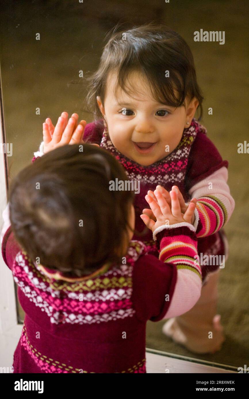 A smiling, brown haired baby girl plays with her reflection in a mirror ...