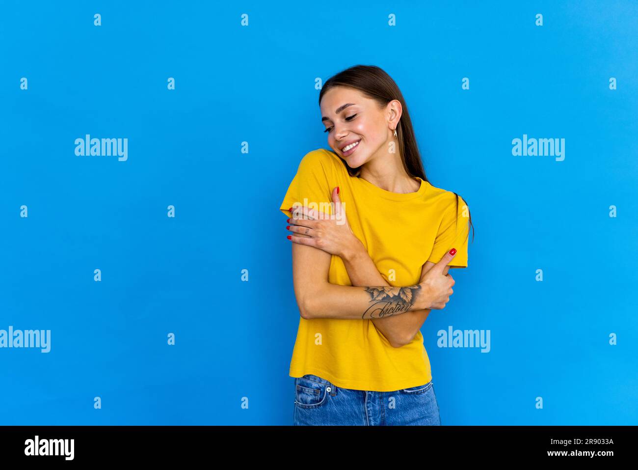 Young woman standing over blue background hugging oneself happy and ...