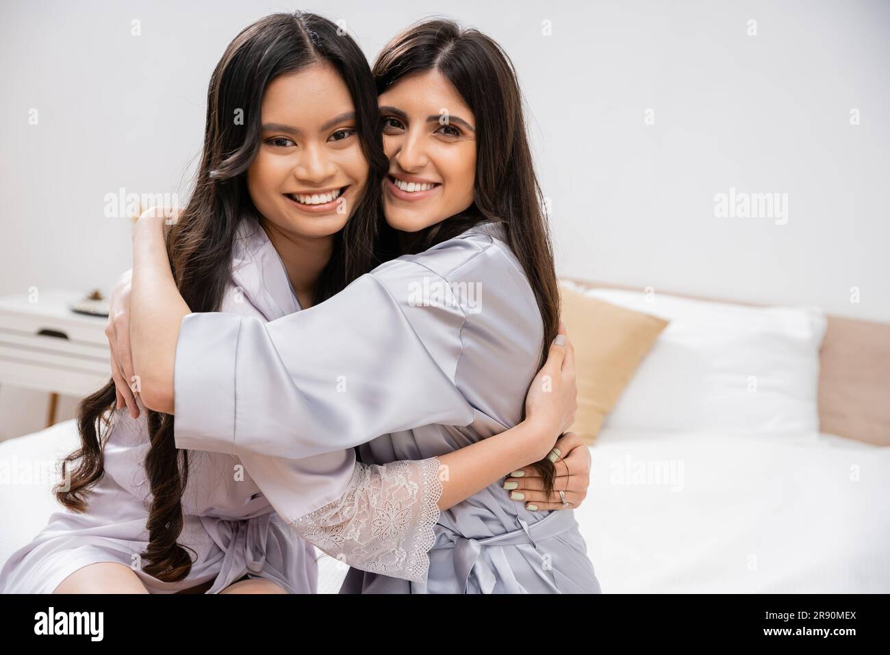 happy asian woman and her friend sitting on bed, hugging each other ...