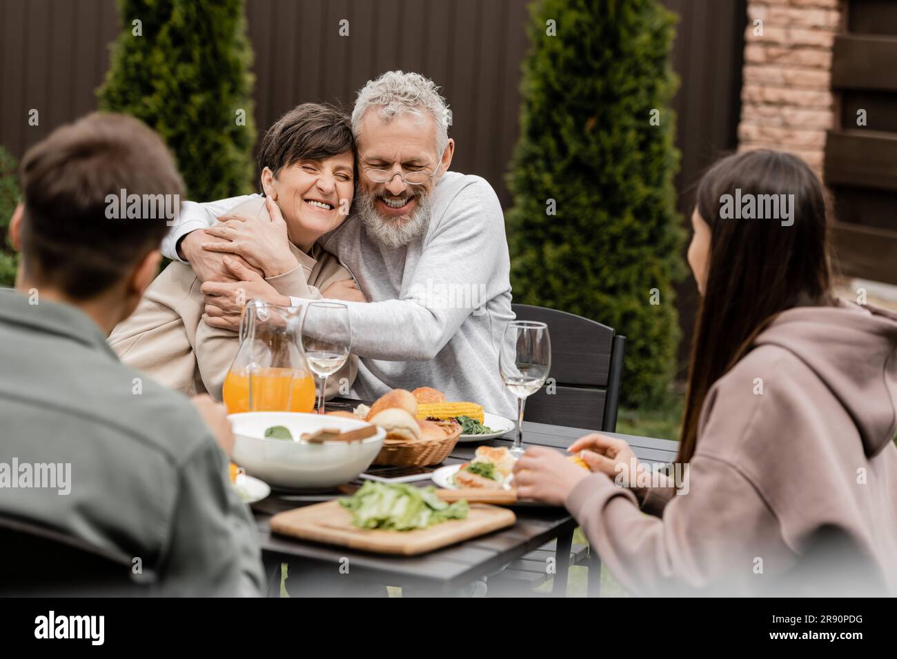 Cheerful middle aged couple hugging near blurred kids and summer food ...