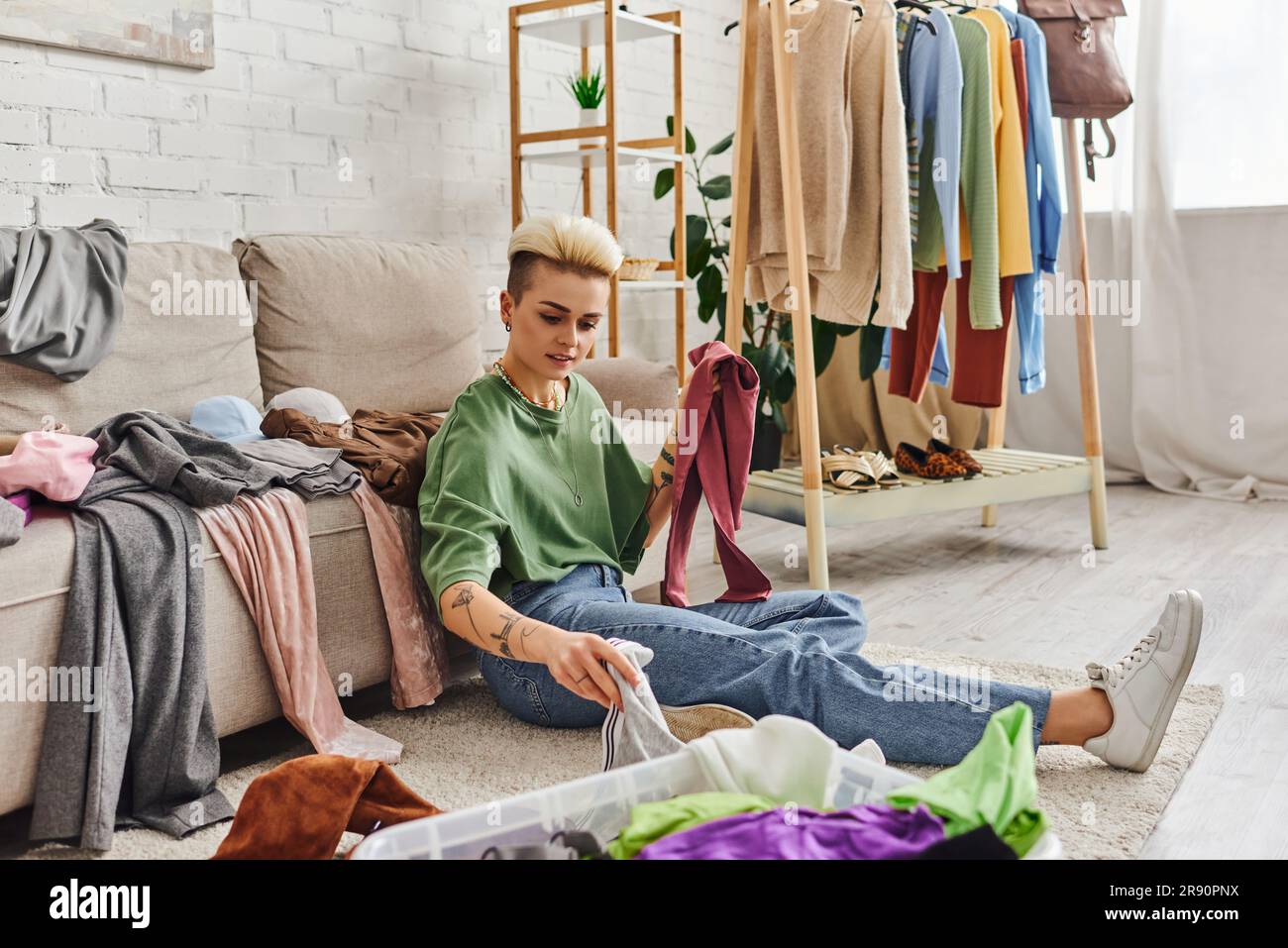 tattooed woman sorting wardrobe items while sitting on floor in living ...