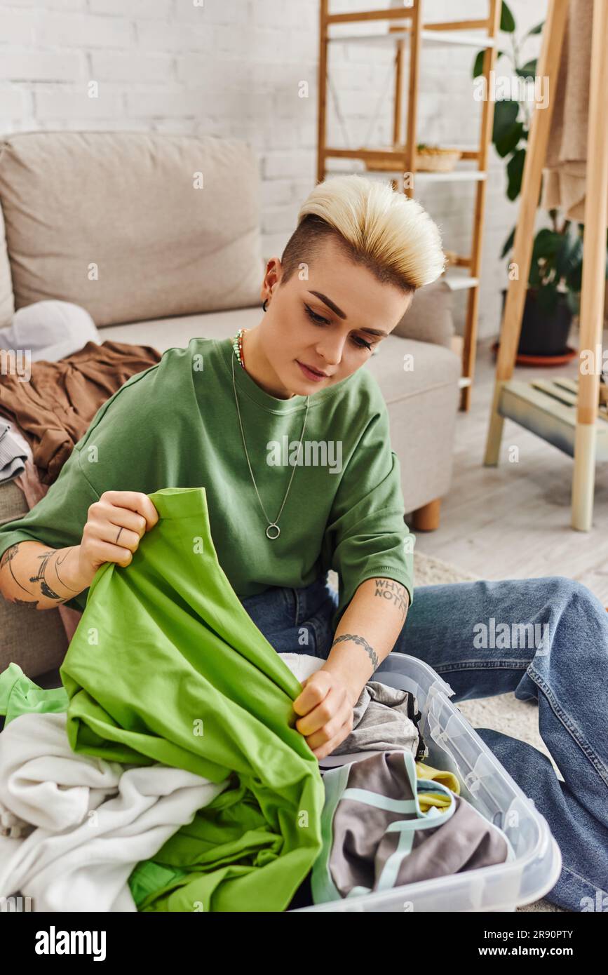 young woman with tattoo and trendy hairstyle holding green garment near ...