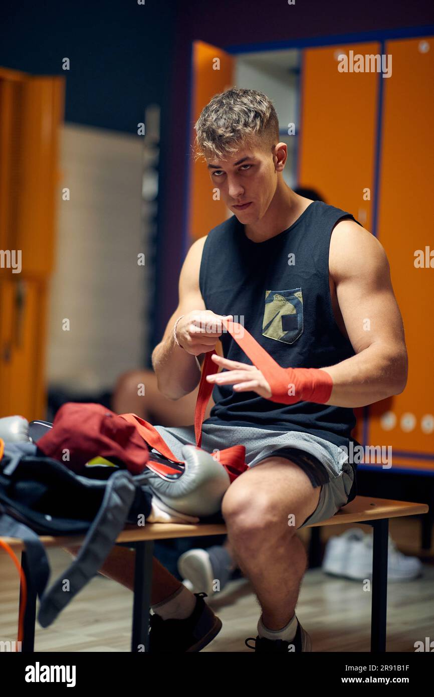 Young man in dressing room, sitting on bench, tying red tape around his ...