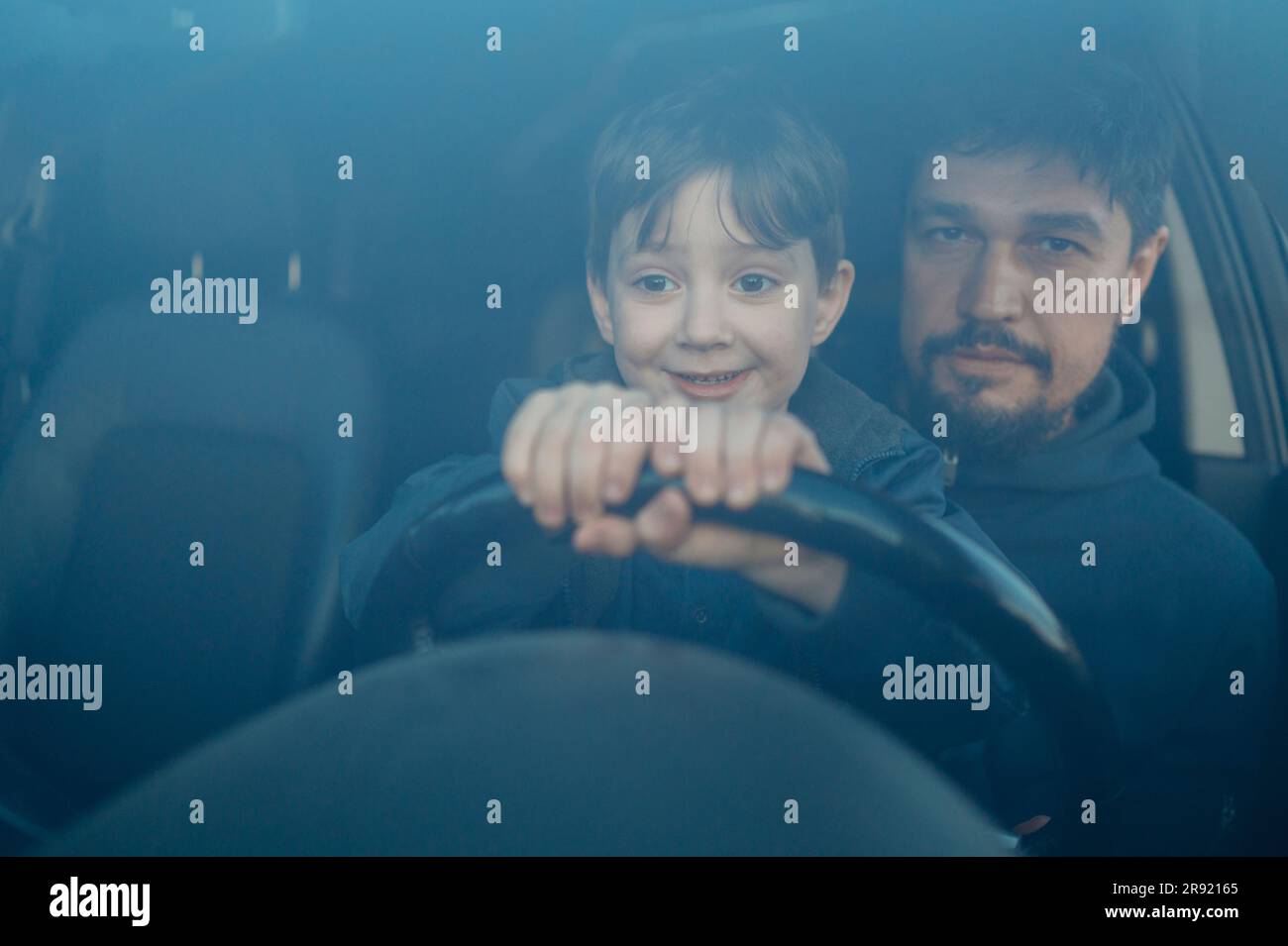 Smiling son learning to drive car with father seen through windshield ...