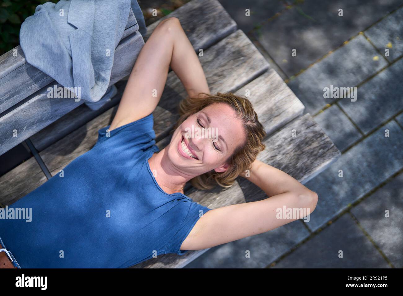 Carefree smiling woman with hands behind head lying on bench at sunny ...