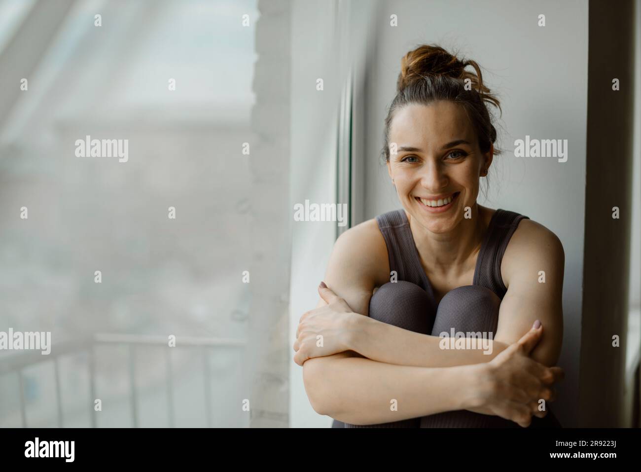 Happy woman hugging knees near window at home Stock Photo - Alamy
