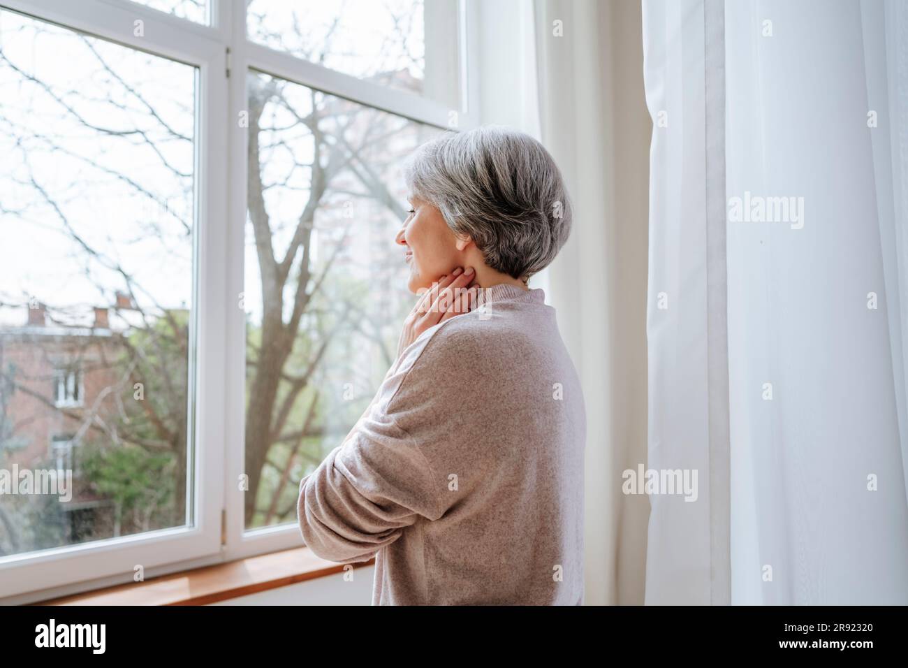 Thoughtful mature woman looking through window at home Stock Photo - Alamy