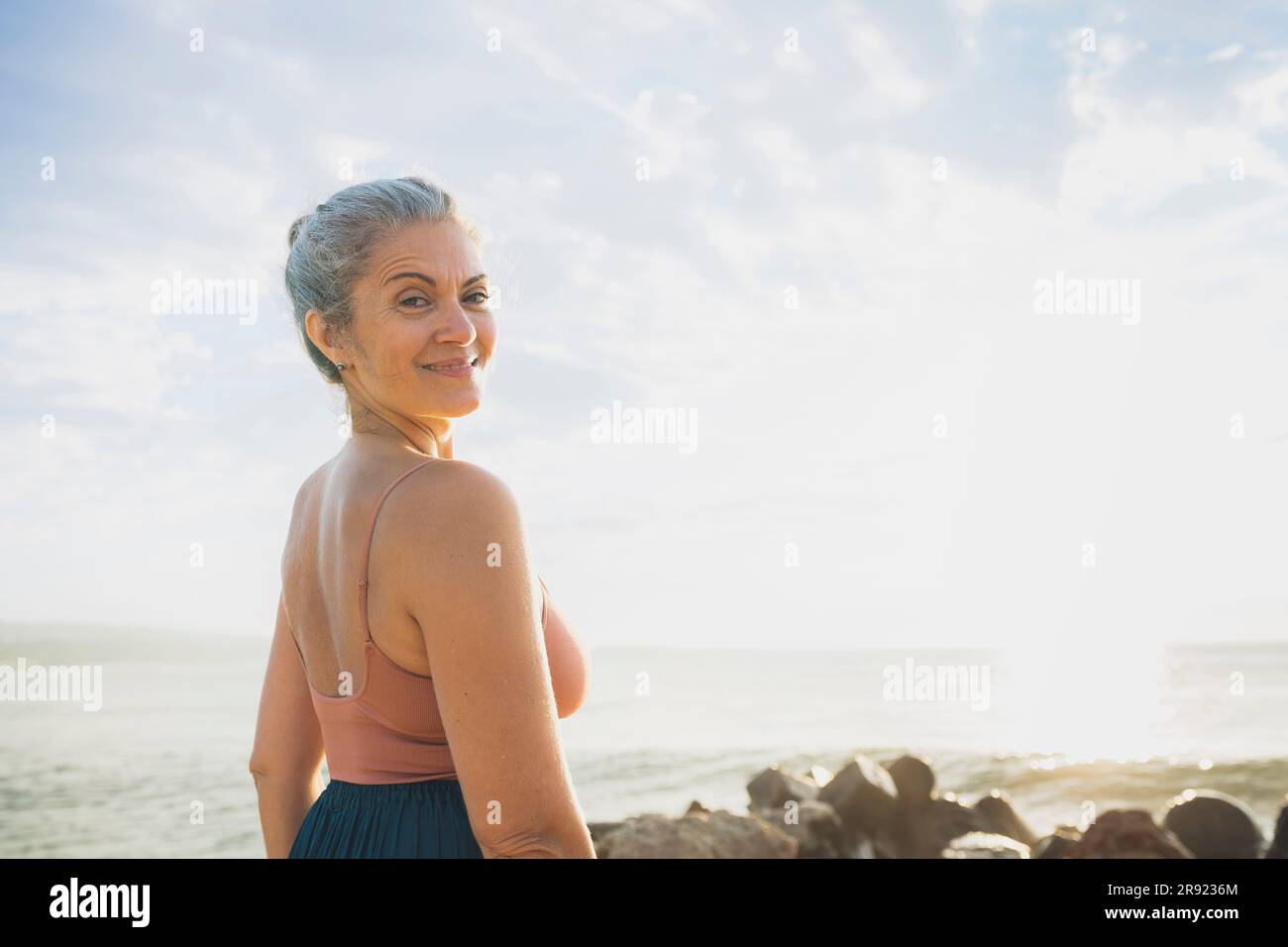 Smiling mature woman looking over shoulder in front of sky at beach ...