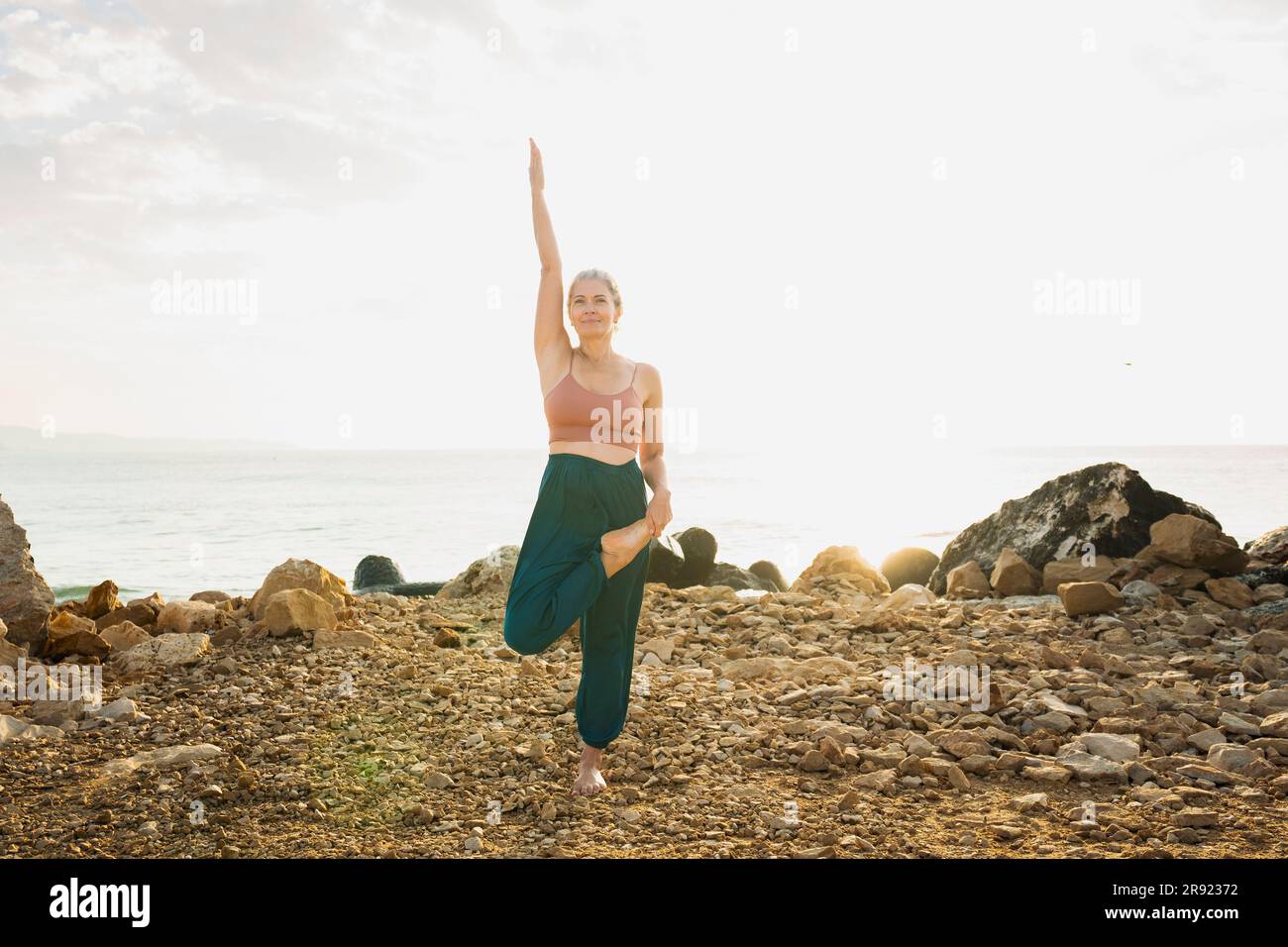 Woman practicing yoga standing on one leg in front of sky at beach ...